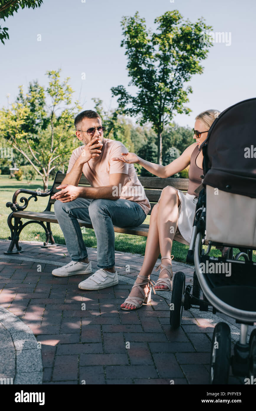 father smoking cigarette near baby carriage in park, angry mother ...