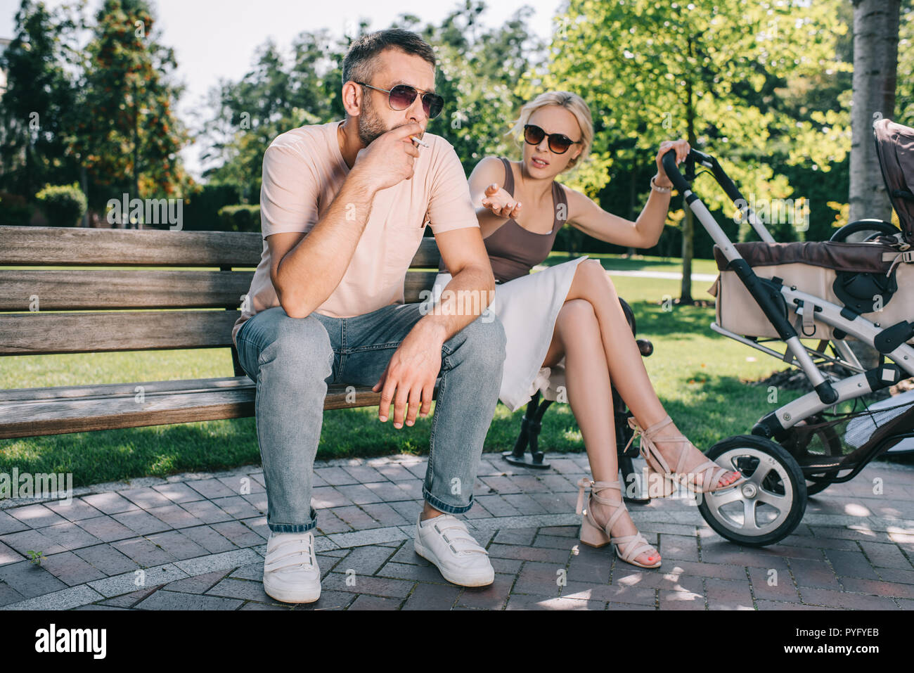 father smoking cigarette near baby carriage in park, mother gesturing ...