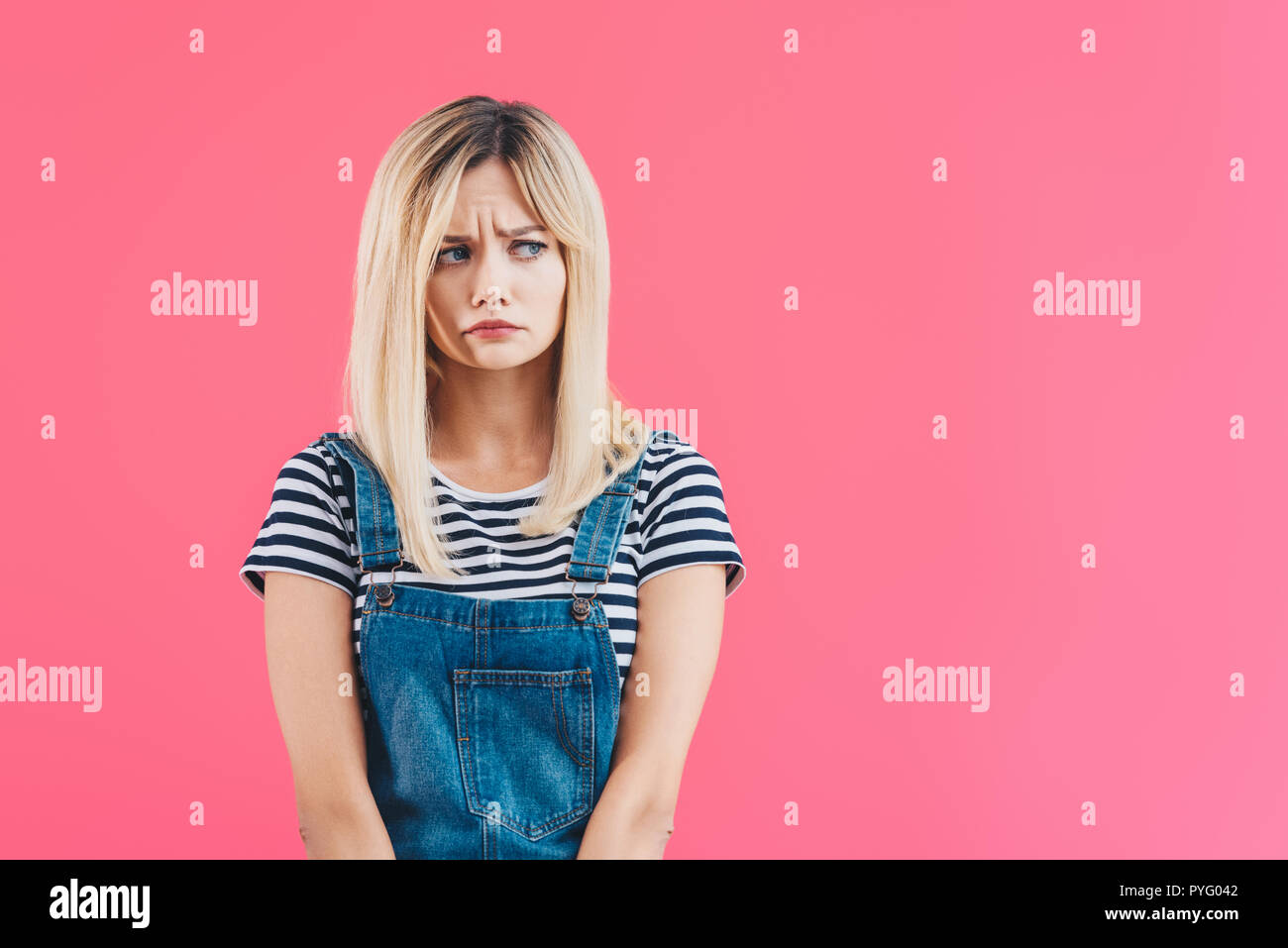 worried beautiful girl in denim overall looking away isolated on pink ...