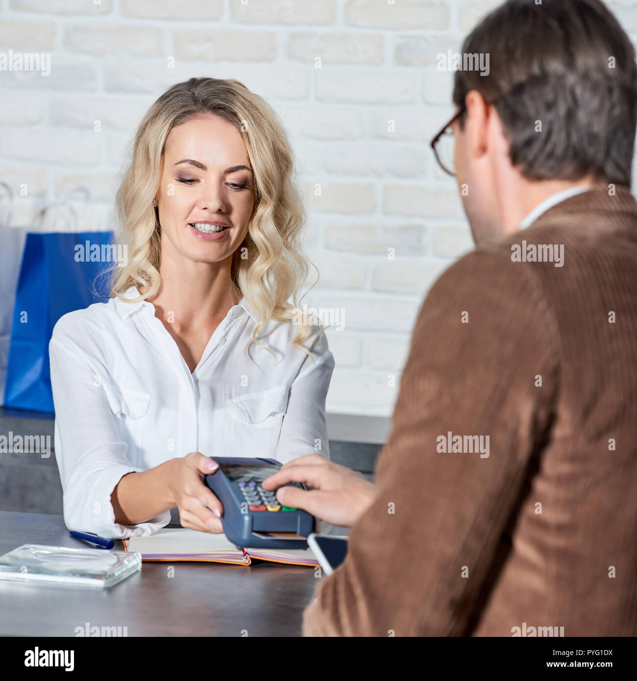 smiling female seller holding payment terminal and customer entering ...