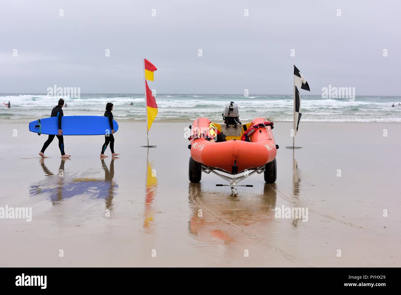 People carrying a blue surf board walking past RNLI beach safety flags ...