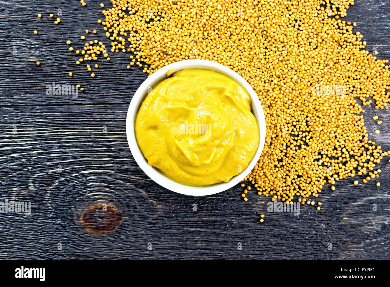 Mustard sauce in a white bowl and seeds on a wooden board background on ...