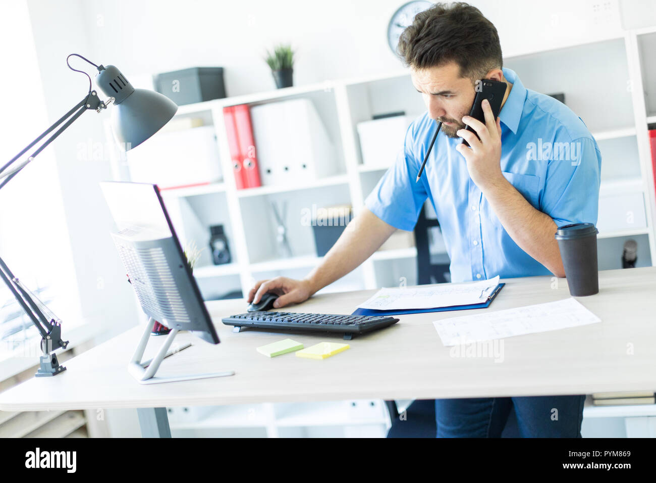 A young man standing in the office at the computer Desk and talking on ...