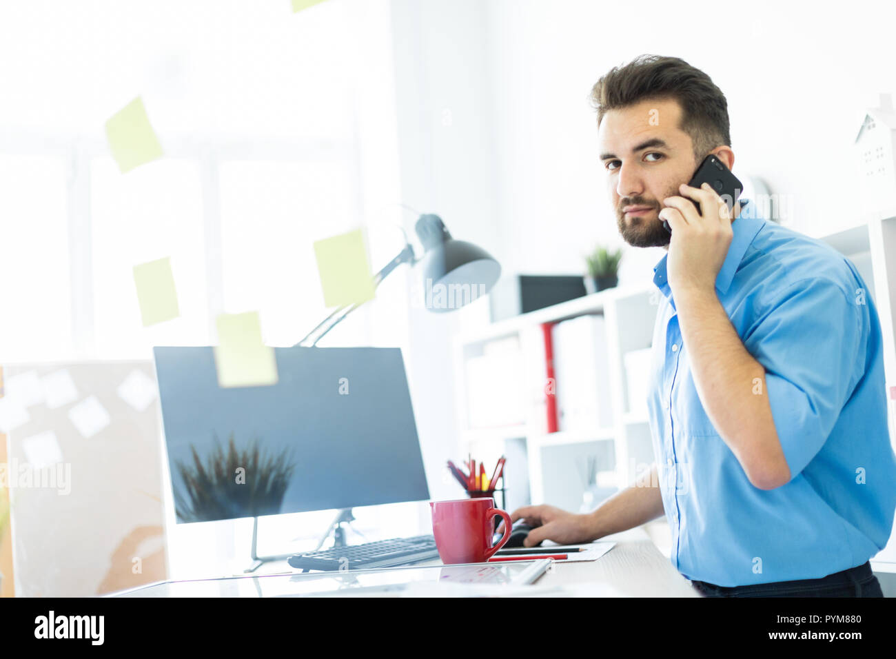 A young man standing in the office at the computer Desk and talking on ...