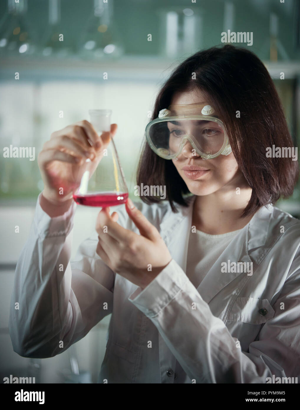 Chemical laboratory. Young woman in work glasses looking at the flask ...