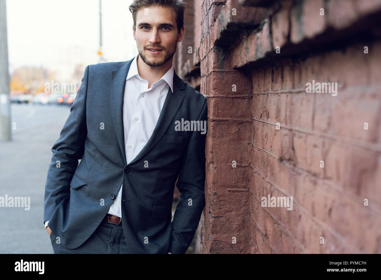 Smiling young male manager formal dressed leaning on a wall outdoors ...