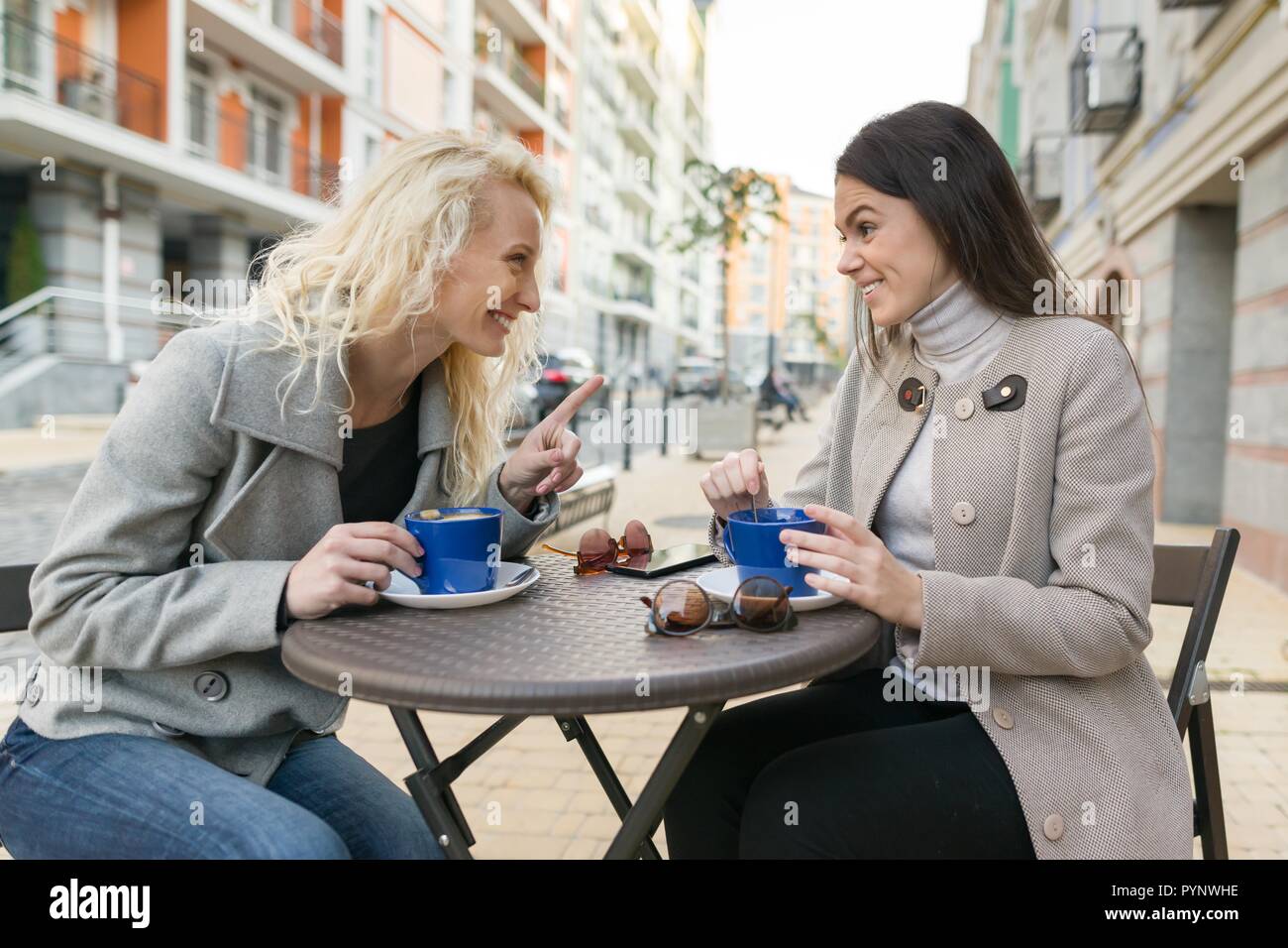 Two young smiling women in an outdoor cafe, drinking coffee, talking ...