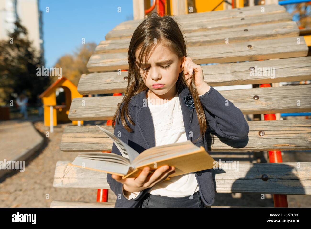 Outdoor portrait of offended little girl. A girl is reading thick book ...