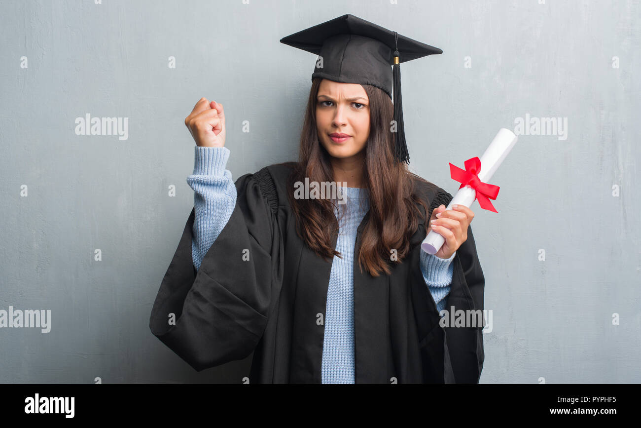 Young brunette woman over grunge grey wall wearing graduate uniform ...
