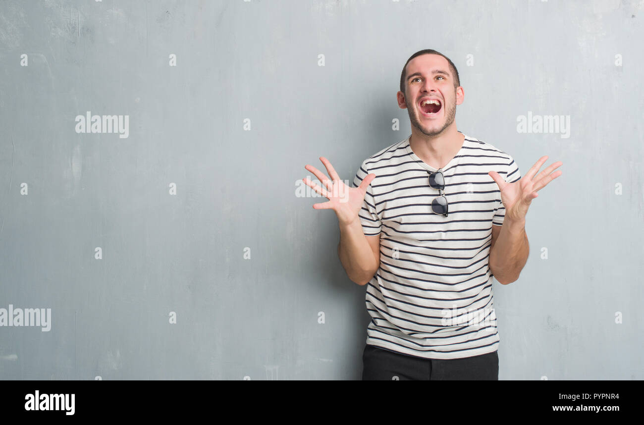 Young caucasian man over grey grunge wall crazy and mad shouting and ...