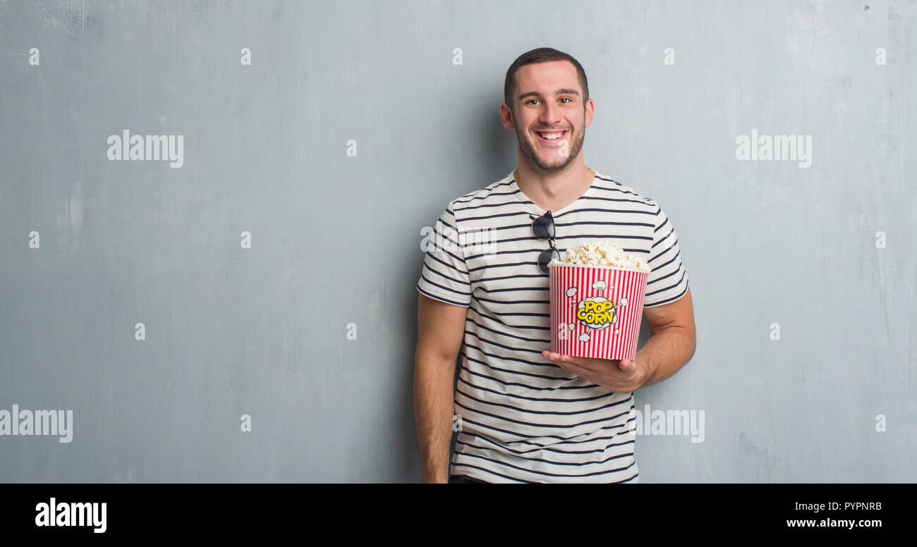 Young caucasian man over grey grunge wall eating popcorn with a happy ...