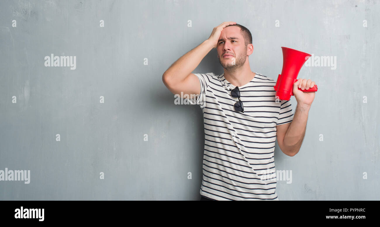 Young caucasian man over grey grunge wall shouting trough megaphone ...