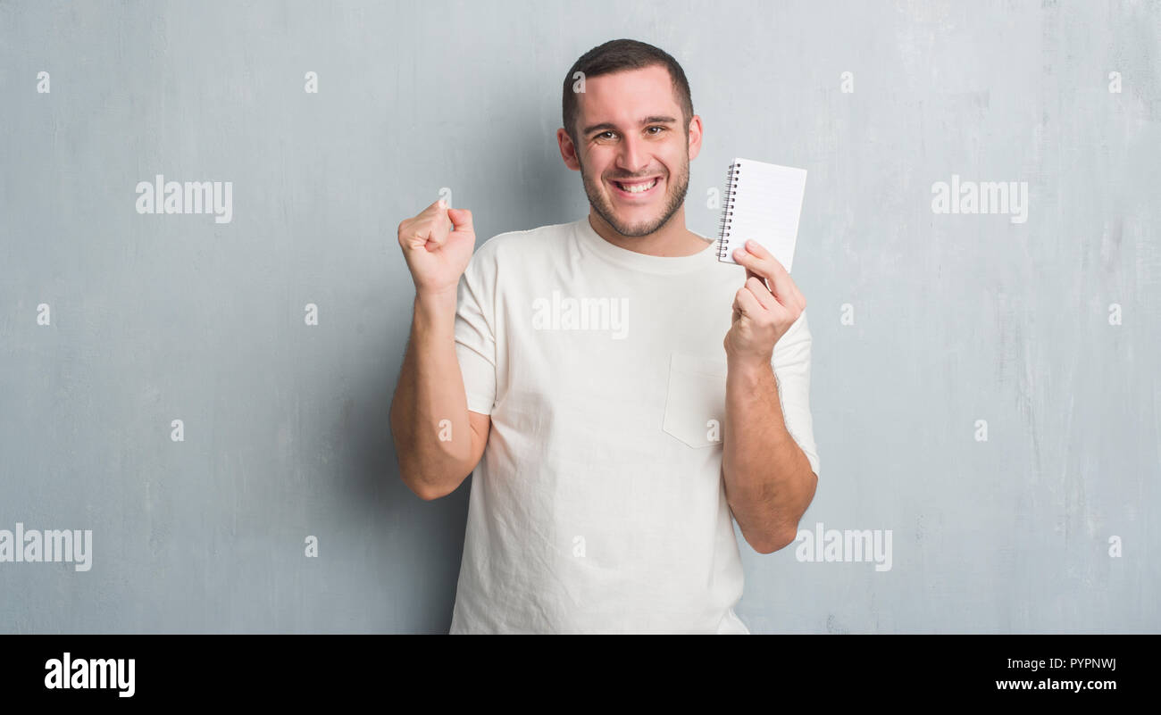 Young caucasian man over grey grunge wall showing notebook screaming ...