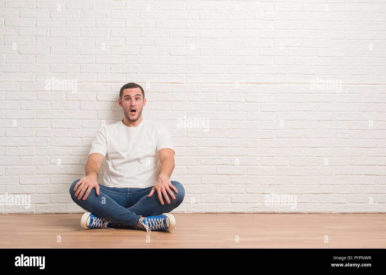 Young caucasian man sitting on the floor over white brick wall afraid ...