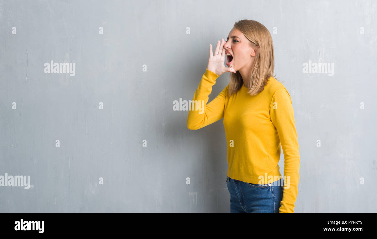 Beautiful young woman standing over grunge grey wall shouting and ...