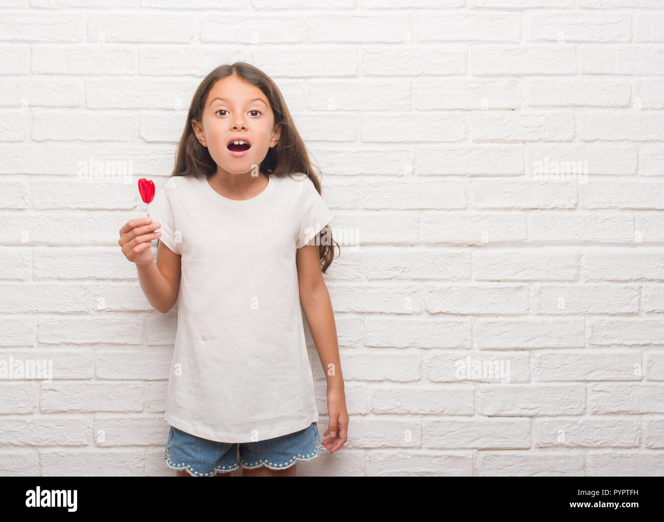 Young hispanic kid over white brick wall eating red heart lollipop ...