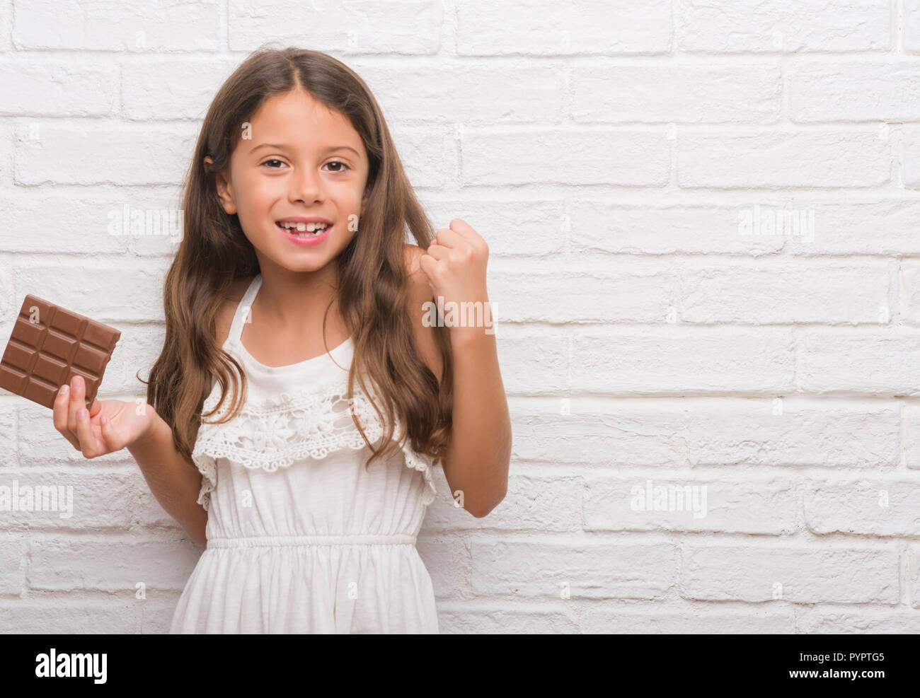 Young hispanic kid over white brick wall eating chocolate bar screaming ...
