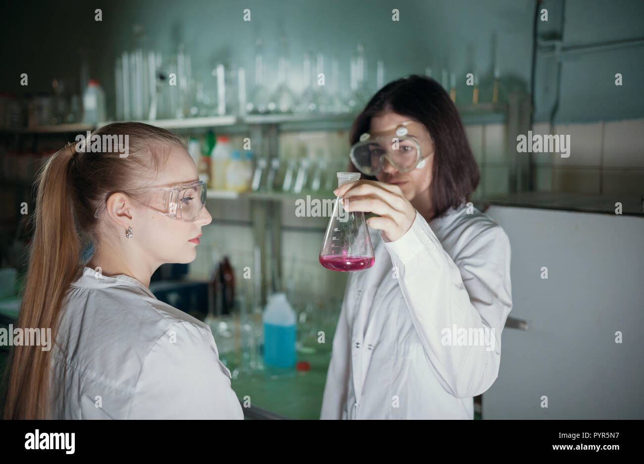 Two young woman in chemical laboratory holding a flask with pink liquid ...