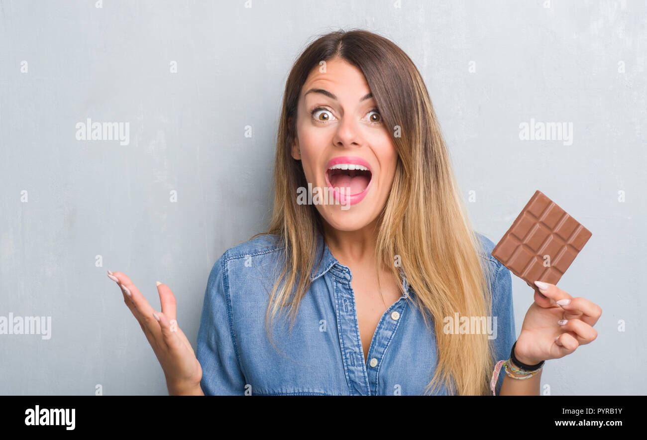 Young adult woman over grey grunge wall eating chocolate bar very happy ...