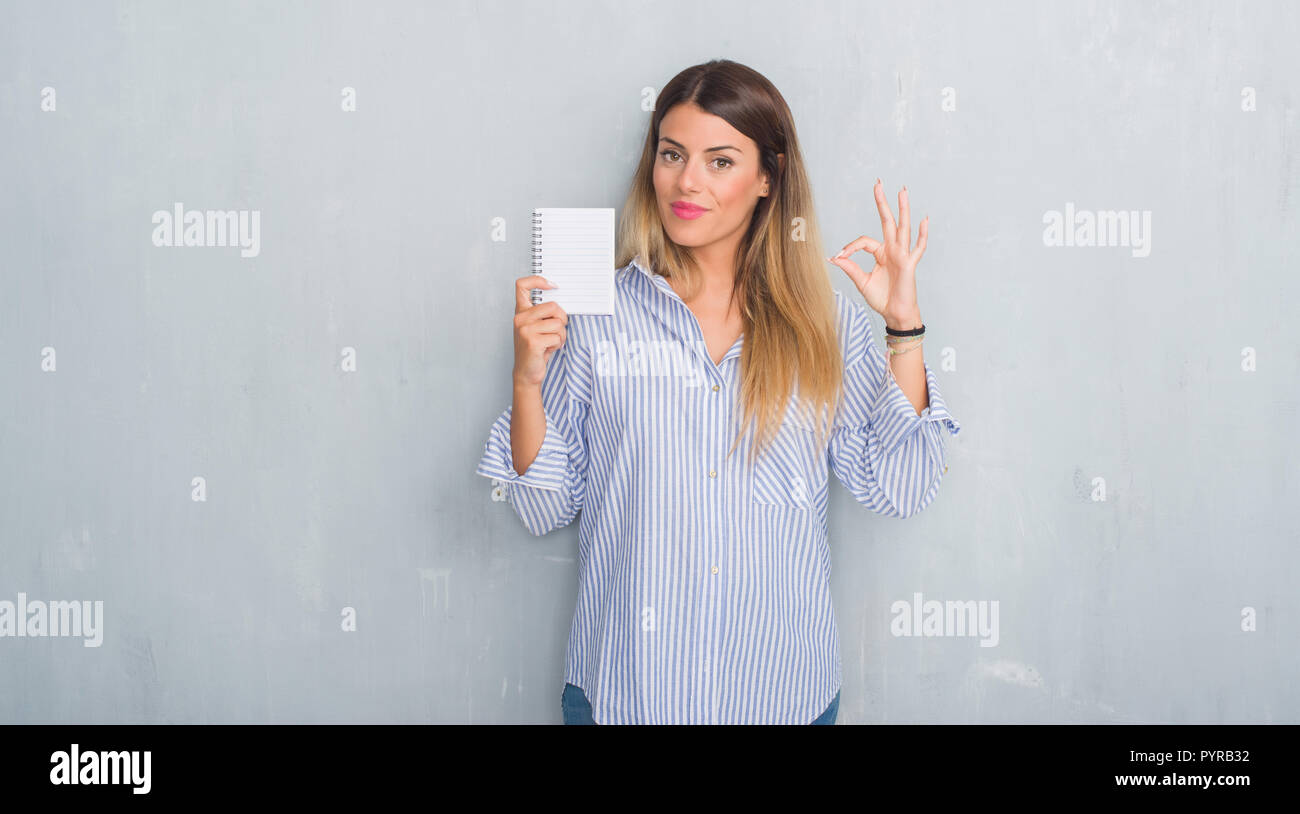 Young adult woman over grey grunge wall holding blank notebook doing ok ...