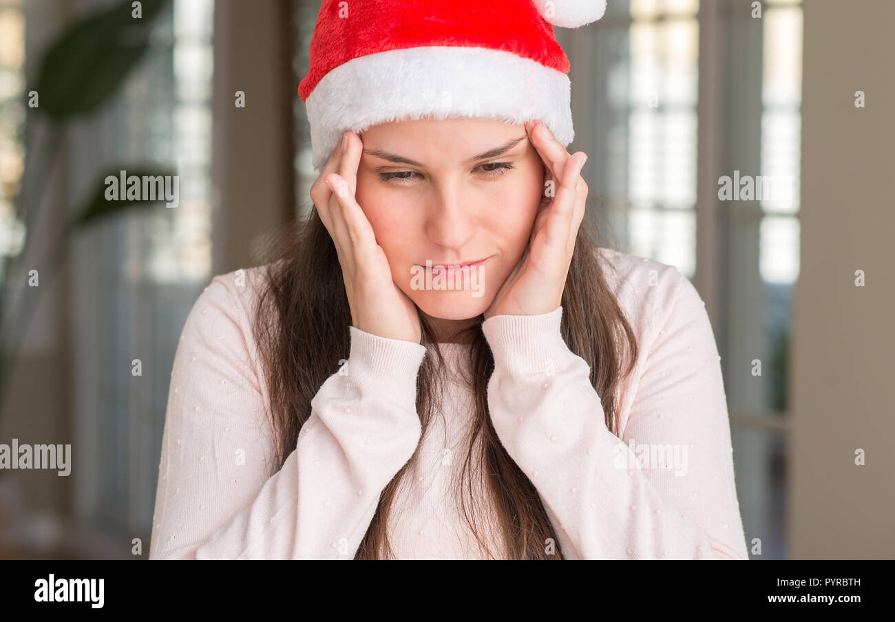 Beautiful young woman wearing Santa Claus hat at home with hand on head ...