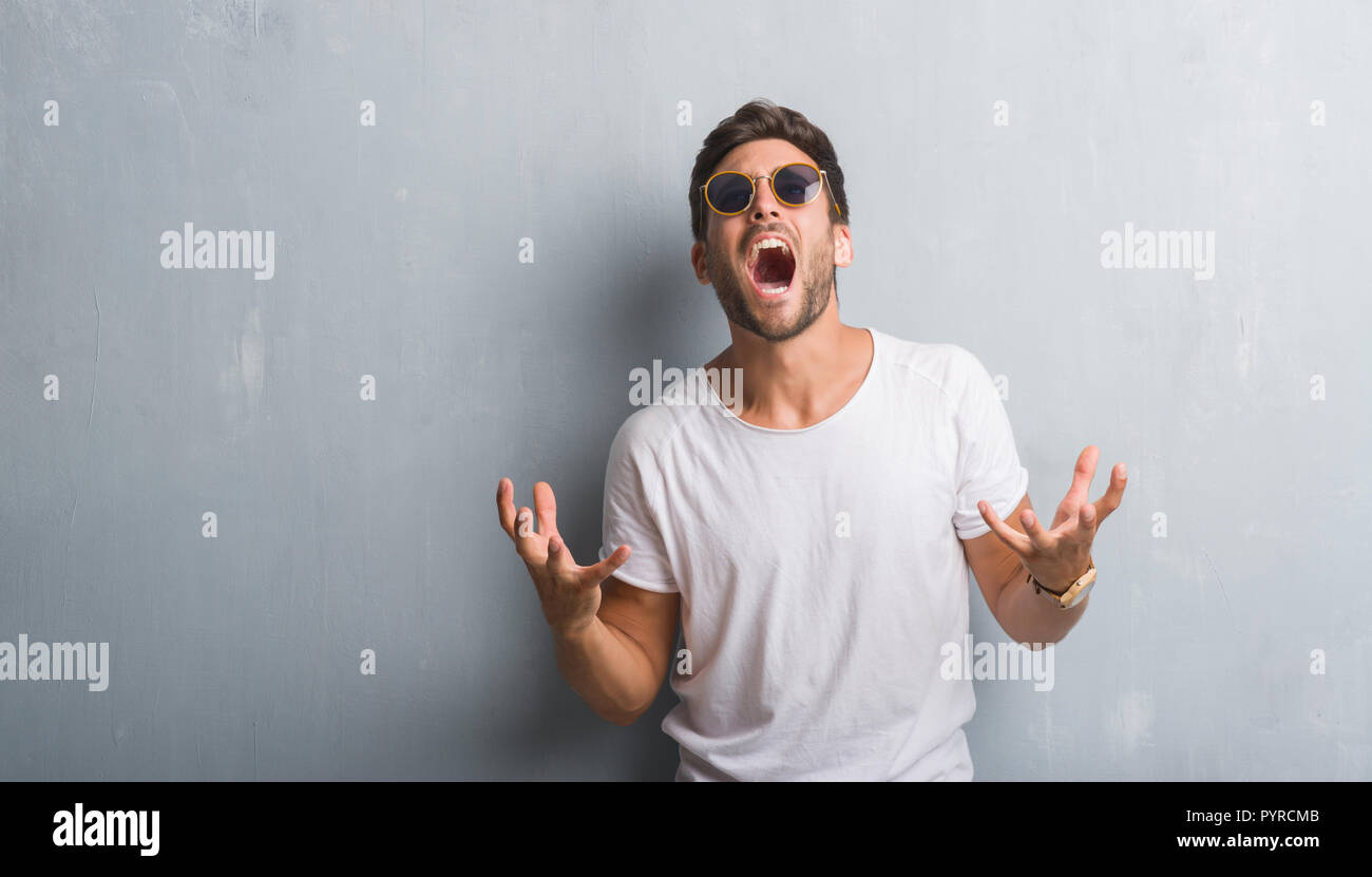 Handsome young man over grey grunge wall wearing sunglasses crazy and ...