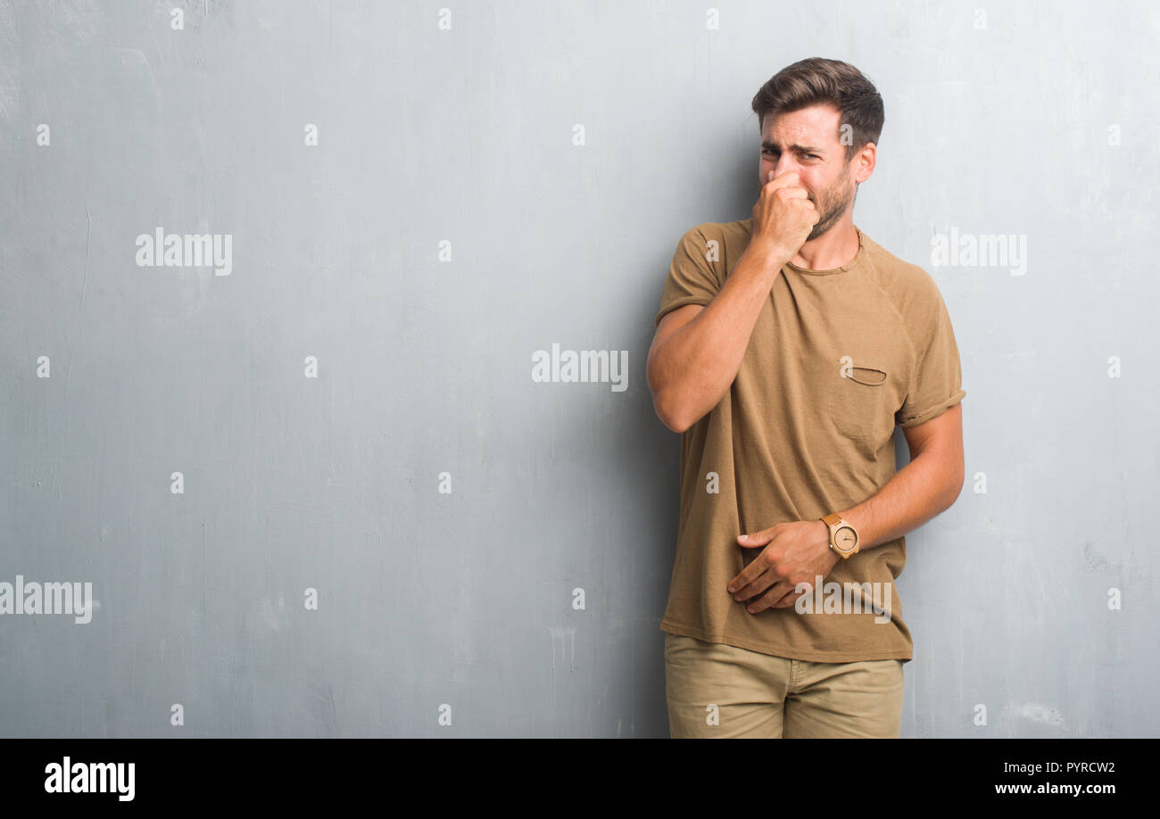 Handsome young man over grey grunge wall smelling something stinky and ...