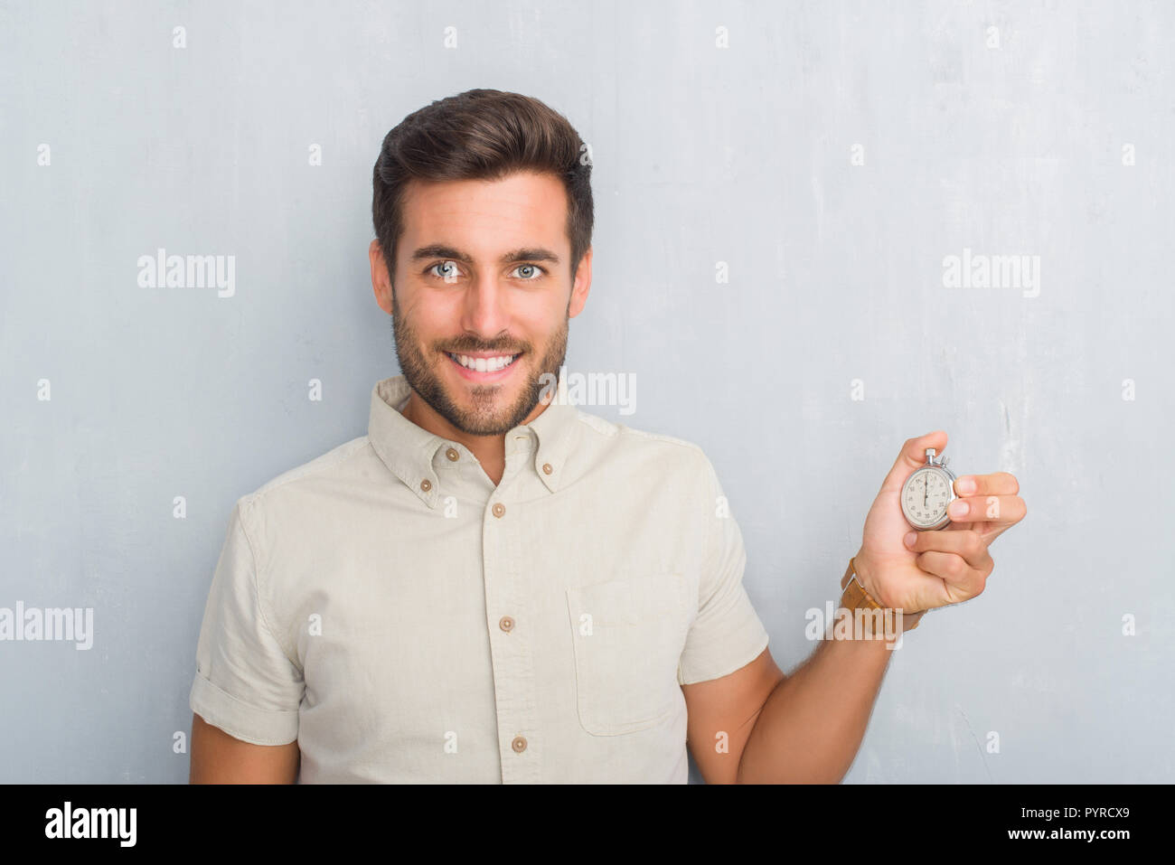 Handsome young man over grey grunge wall holding stopwatch with a happy ...