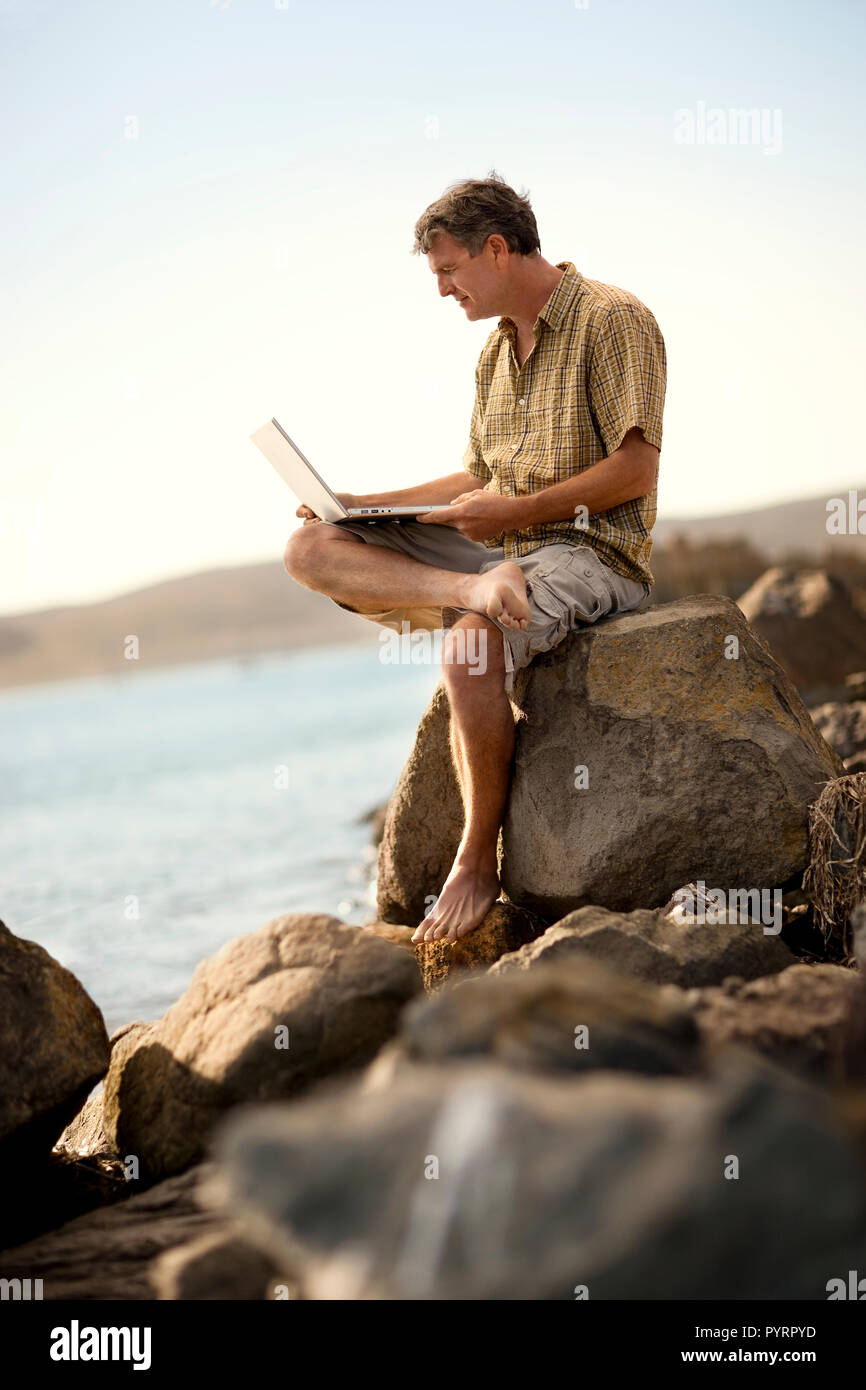 Thoughtful middle aged man sitting on a rock with a laptop computer ...