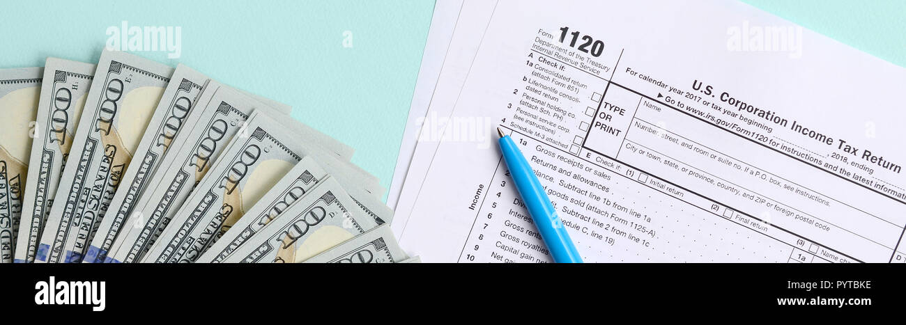 1120 tax form lies near hundred dollar bills and blue pen on a light blue background. US ...