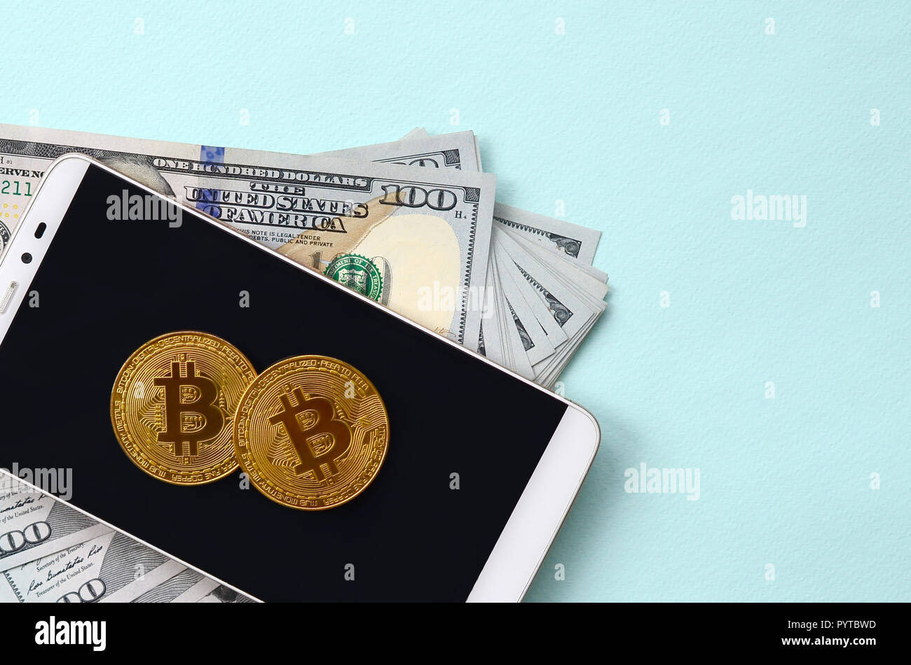 Bitcoins lies on a smartphone and hundred dollar bills on a light blue ...