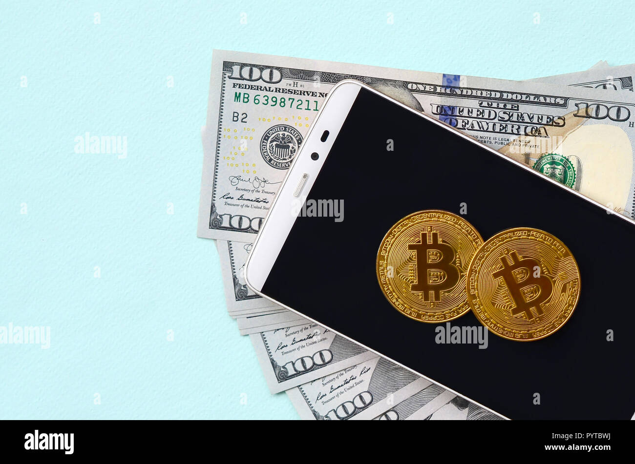 Bitcoins lies on a smartphone and hundred dollar bills on a light blue ...