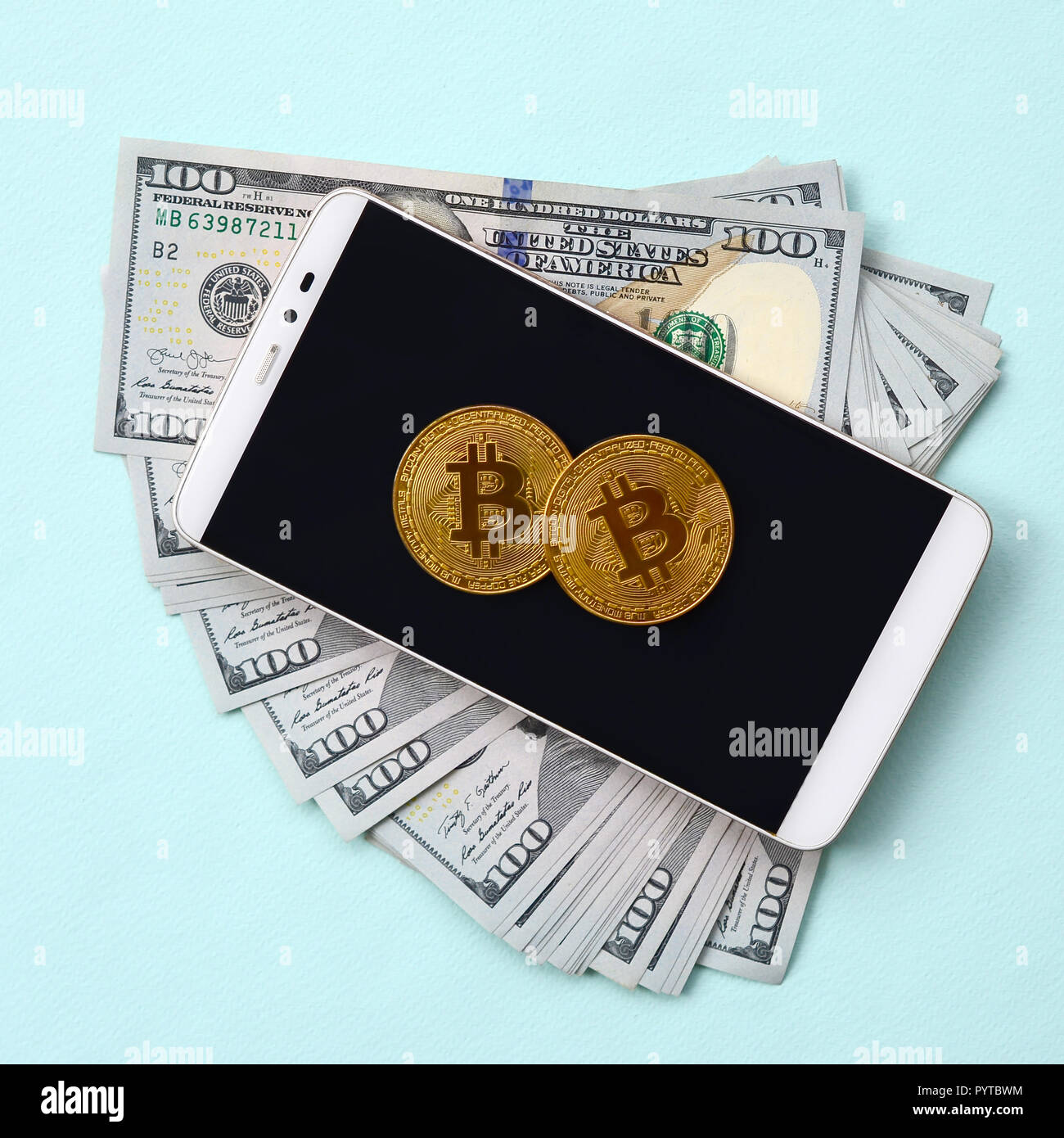 Bitcoins lies on a smartphone and hundred dollar bills on a light blue ...