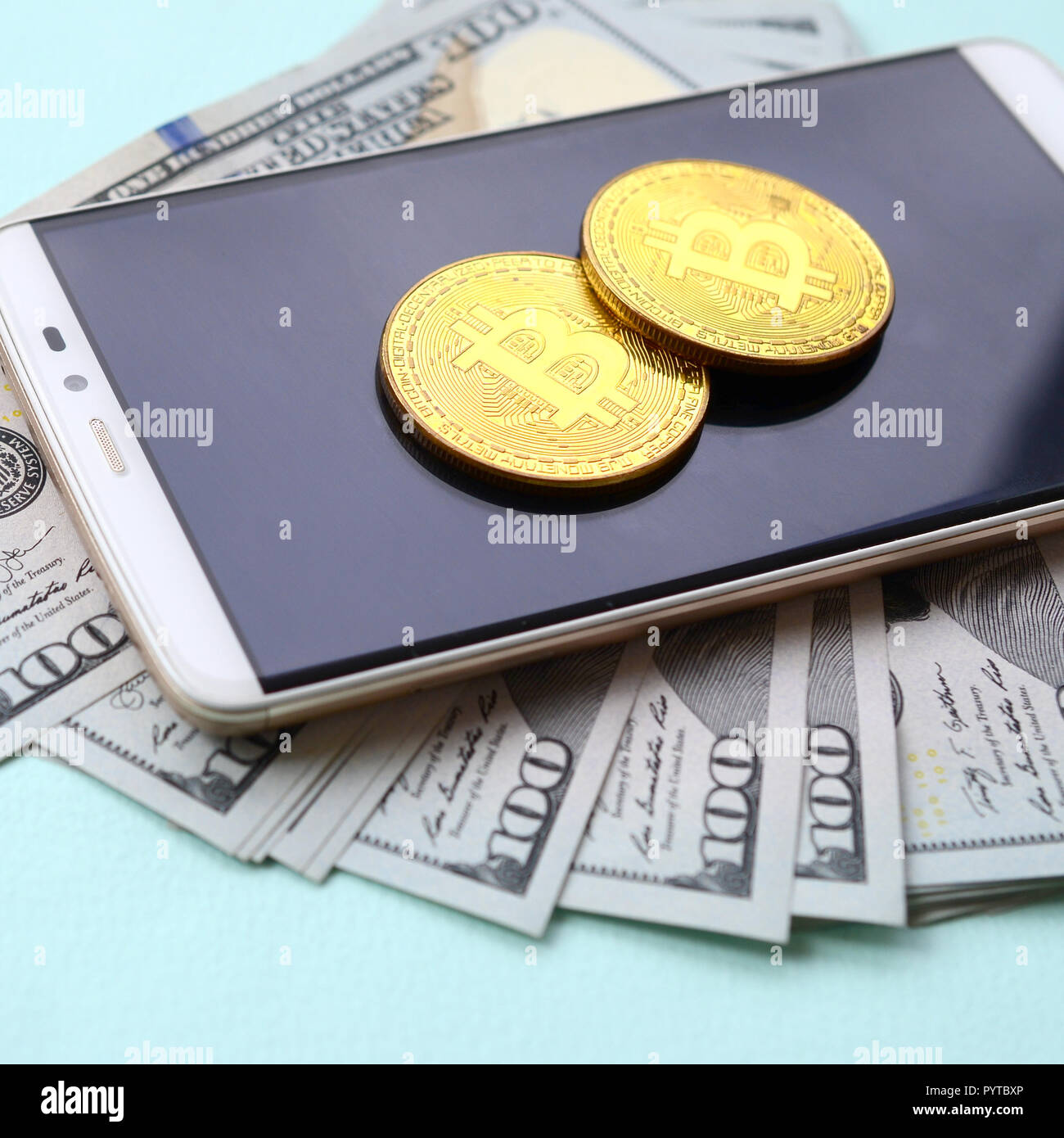 Bitcoins lies on a smartphone and hundred dollar bills on a light blue ...