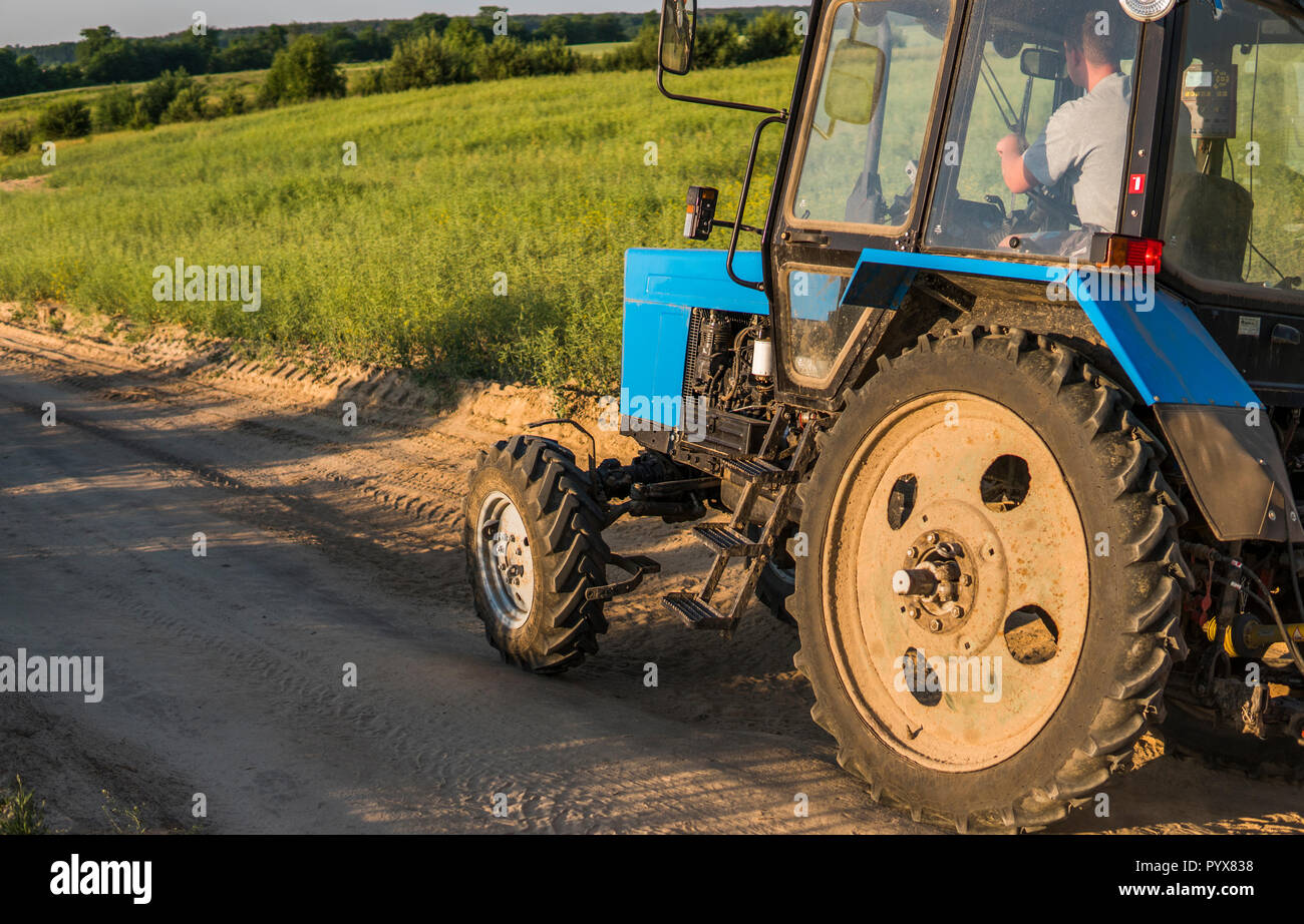 A blue tractor with a trailer for agricultural works rides along a road ...