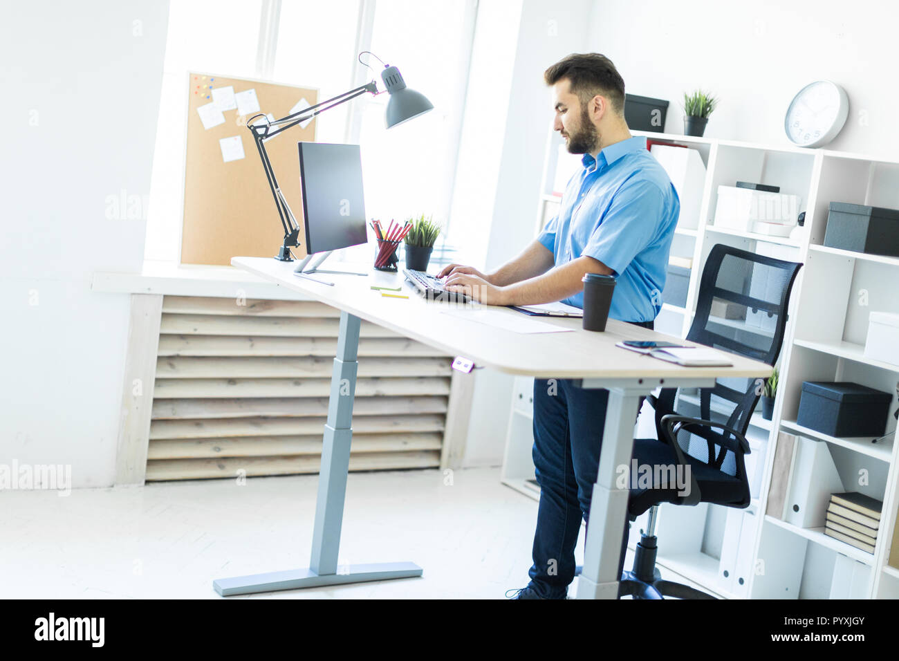 A young man standing in the office and working at the computer table ...