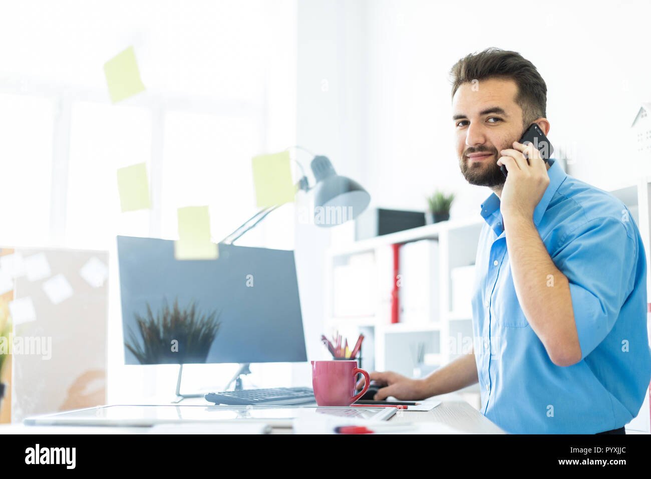 A young man standing in the office at the computer Desk and talking on ...