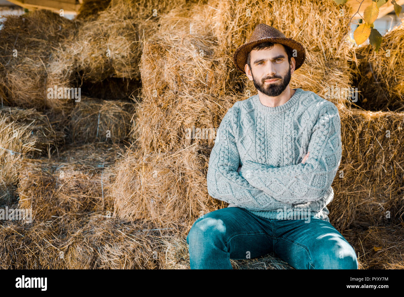 confident male farmer in sweater and straw hat sitting with crossed ...