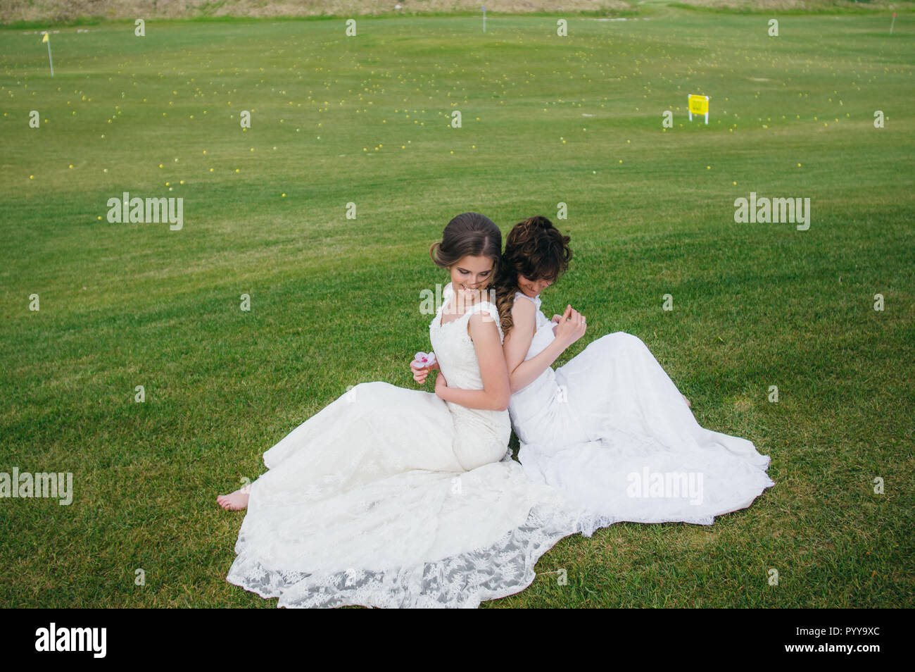 Two beautiful brides sitting back to back on the green field of the ...