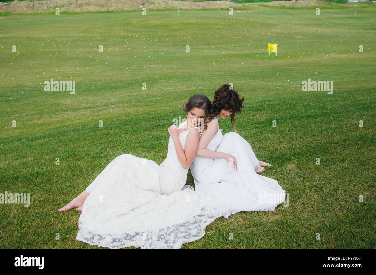 Two beautiful brides sitting back to back on the green field of the ...