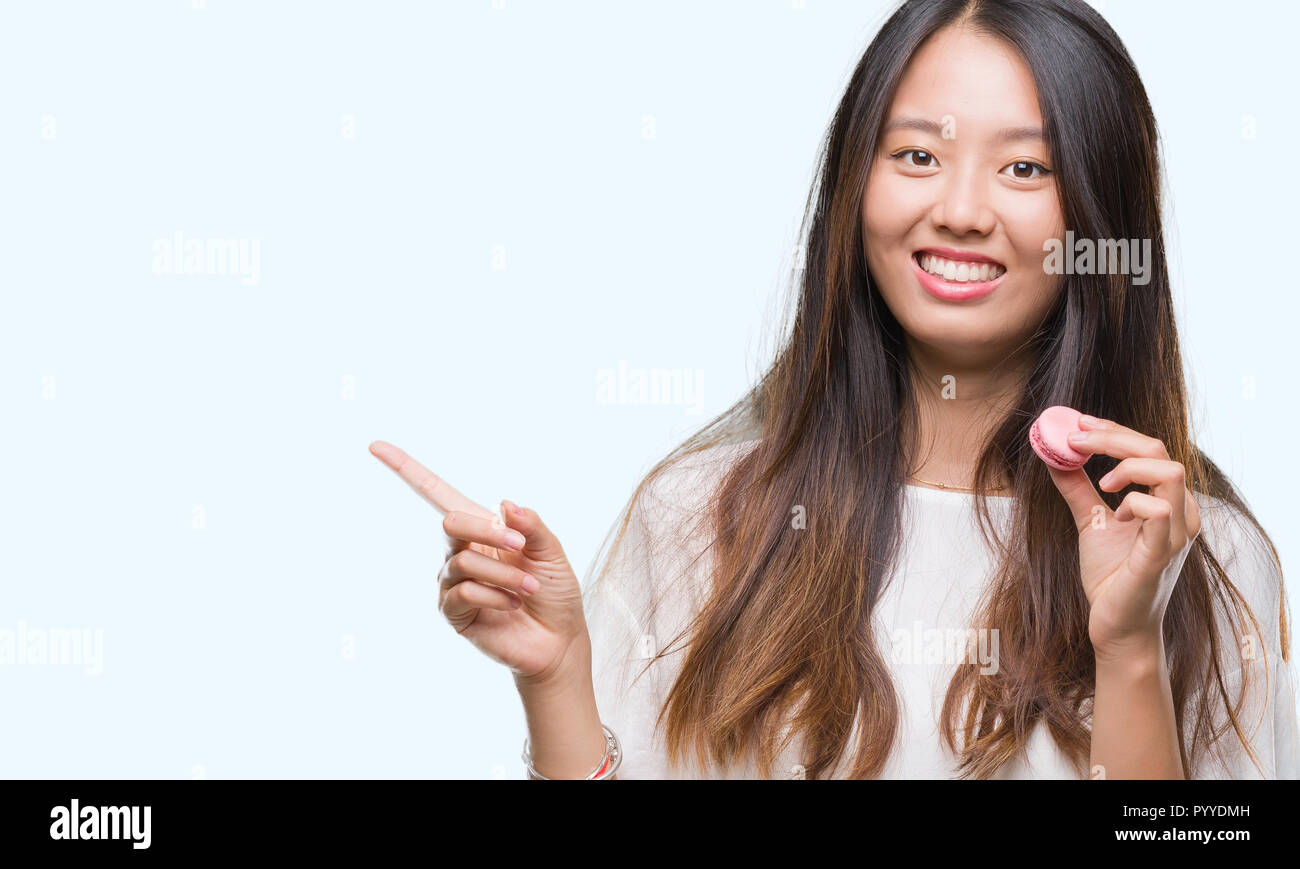 Young asian woman eating pink macaron sweet over isolated background ...