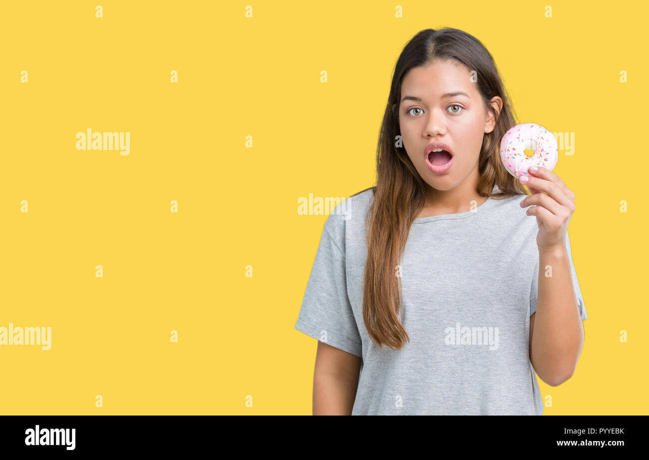 Young beautiful woman eating pink donut over isolated background scared ...