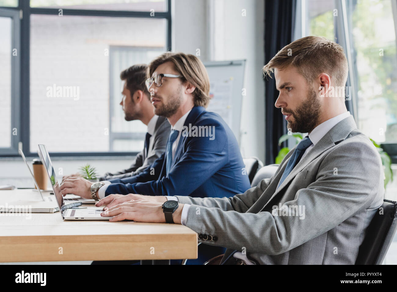 side view of three focused young businessmen using laptops at workplace ...