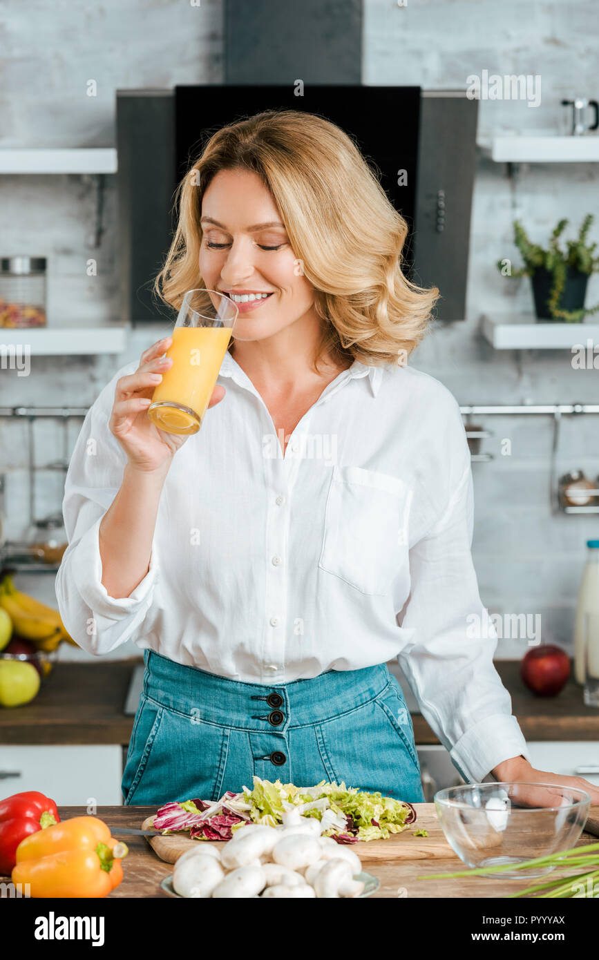 beautiful adult woman drinking orange juice while cooking at kitchen ...