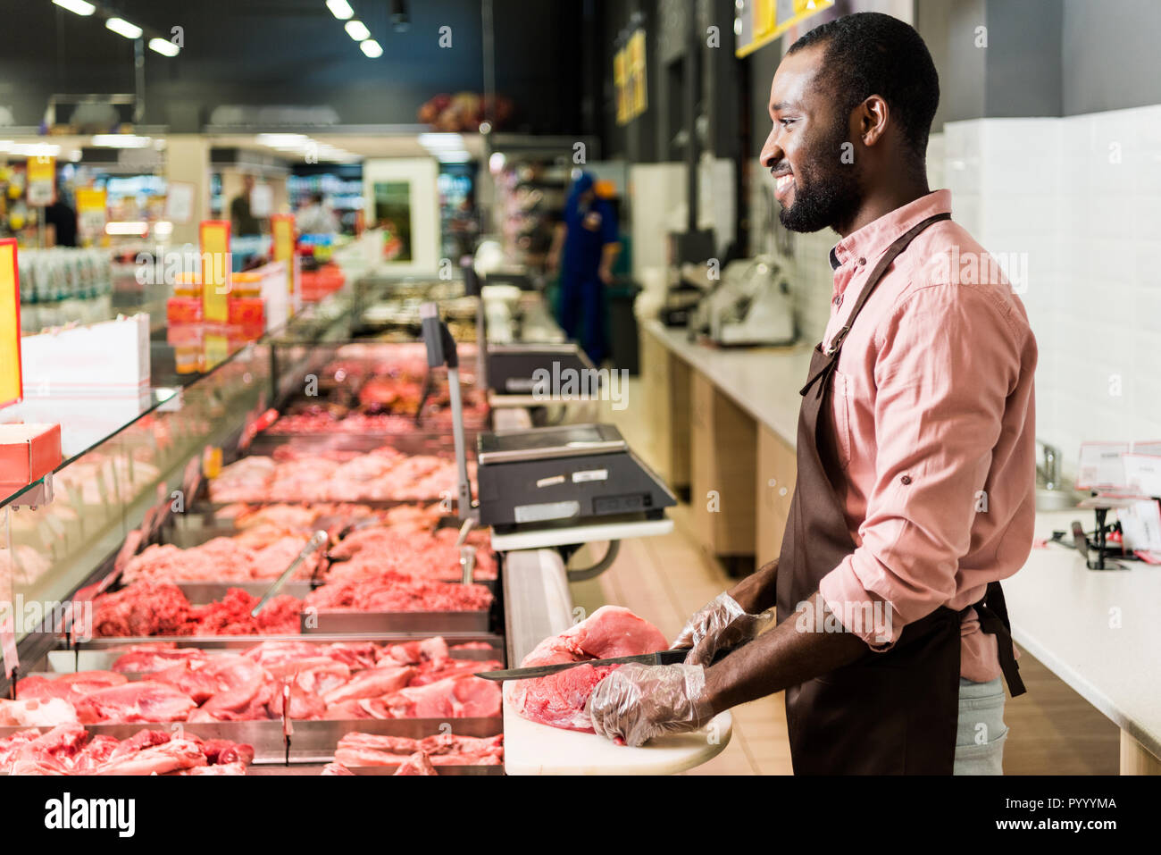 side view of smilng african american male butcher in apron cutting raw ...