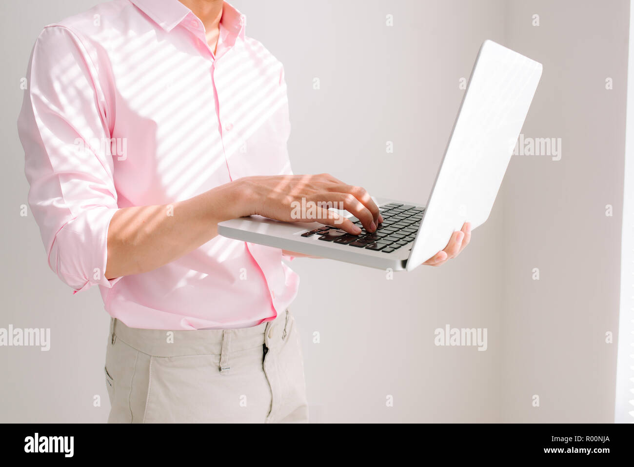 Smiling young man holding laptop and typing while standing against grey ...