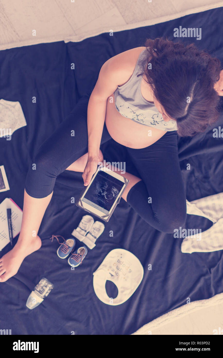 a pregnant woman at home on the floor checking list of baby clothes ...