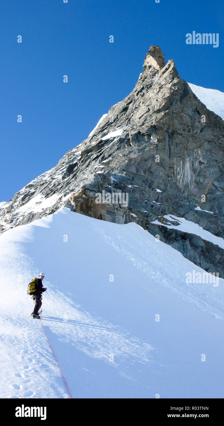 mountain climber standing on a snow and ice ridge below a sharp rock ...