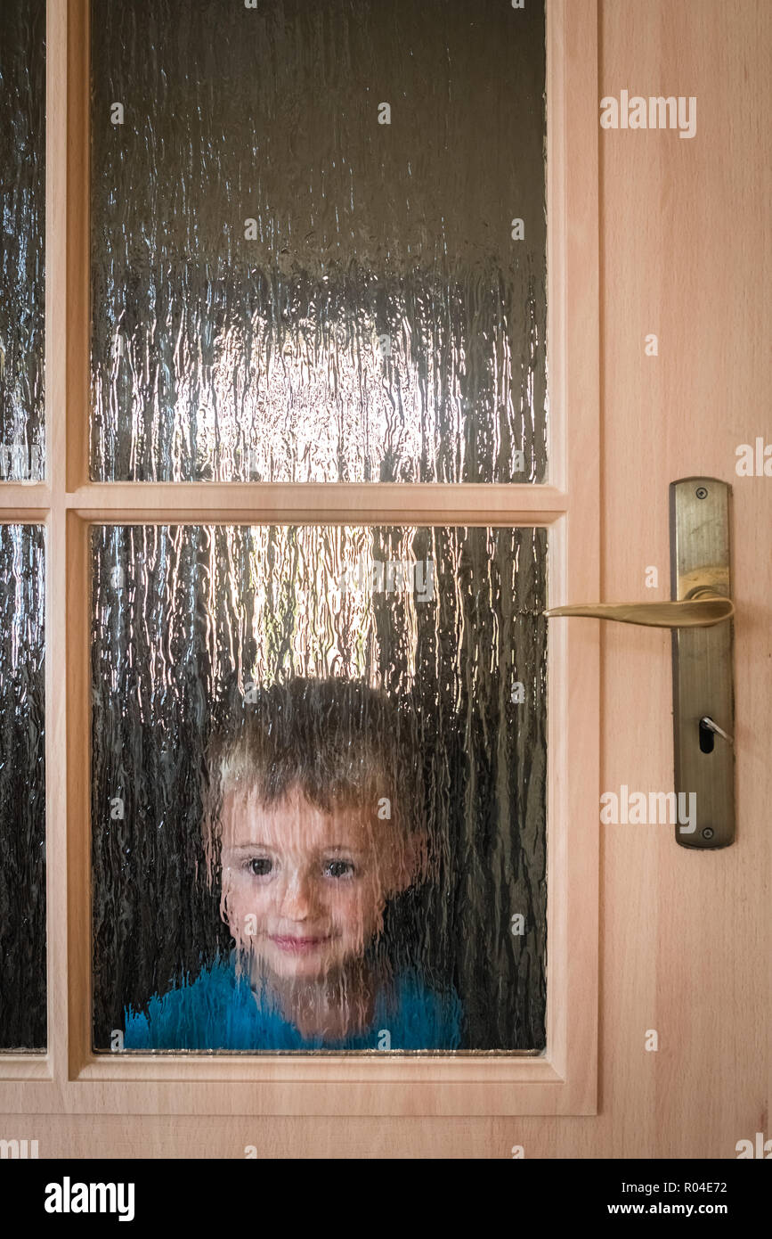 Portrait of a cute little Caucasian boy hiding behind a door with glass ...