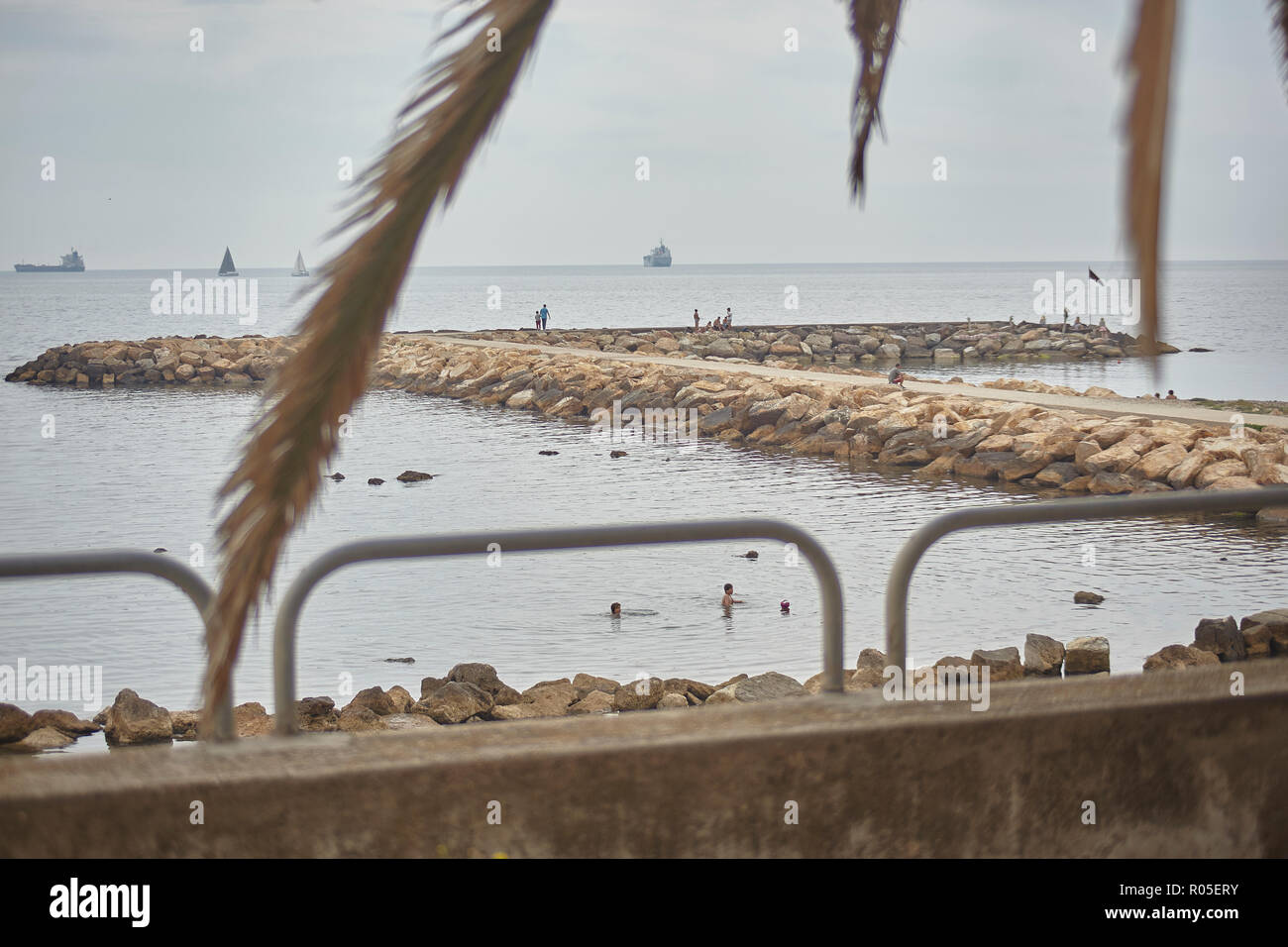 Pier of rocks on the Mediterranean sea filtered by a parapet with ships ...